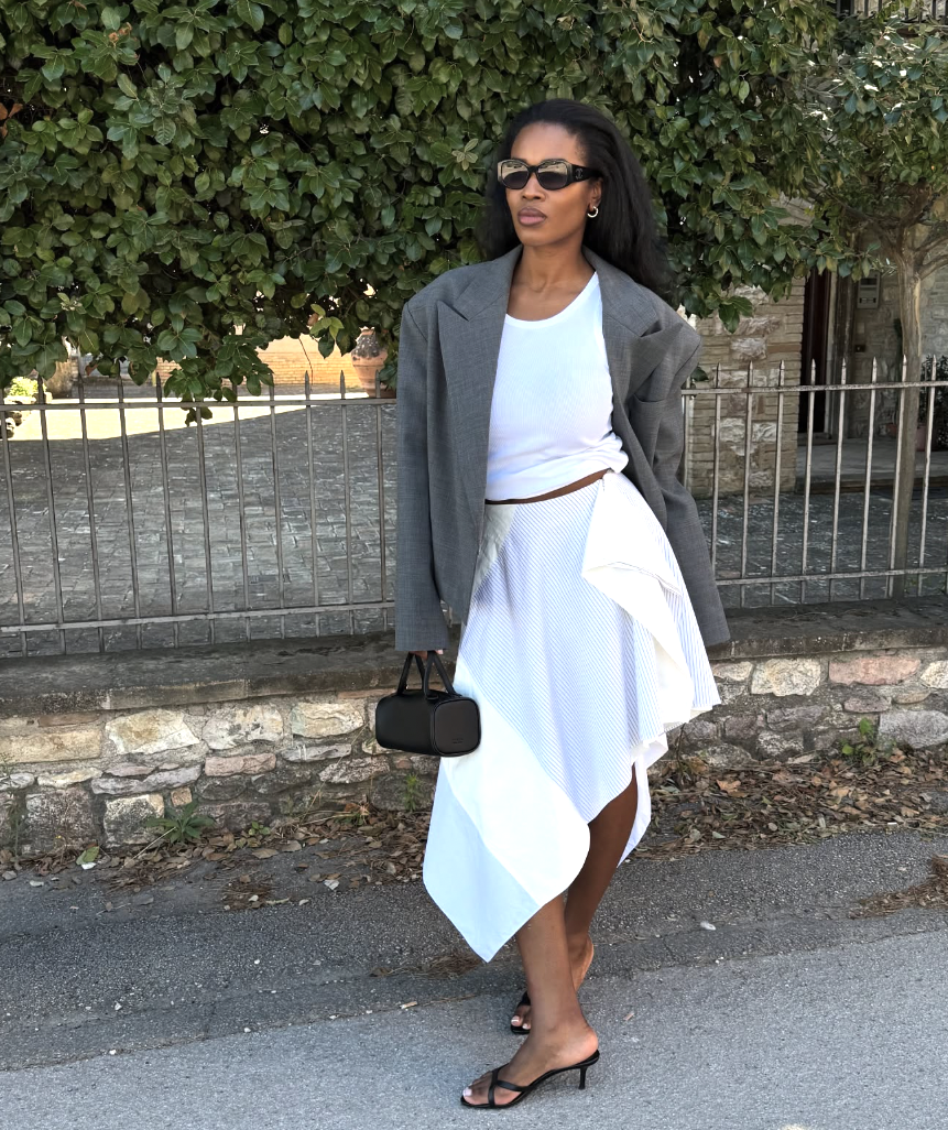 A woman in sunglasses stands confidently in front of a leafy backdrop. She wears a gray blazer%2C white top%2C and asymmetrical skirt%2C holding a small black bag.